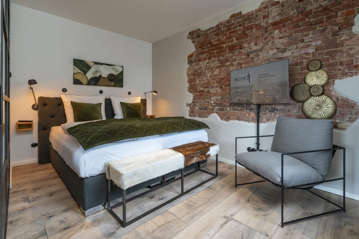 Quarto moderno com parede de tijolos, cama com manta verde, cadeira cinza e placa na parede.