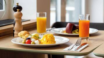 Mesa de café da manhã com ovos mexidos, tomates, azeitonas e um croissant, acompanhada de dois copos de suco de laranja.
