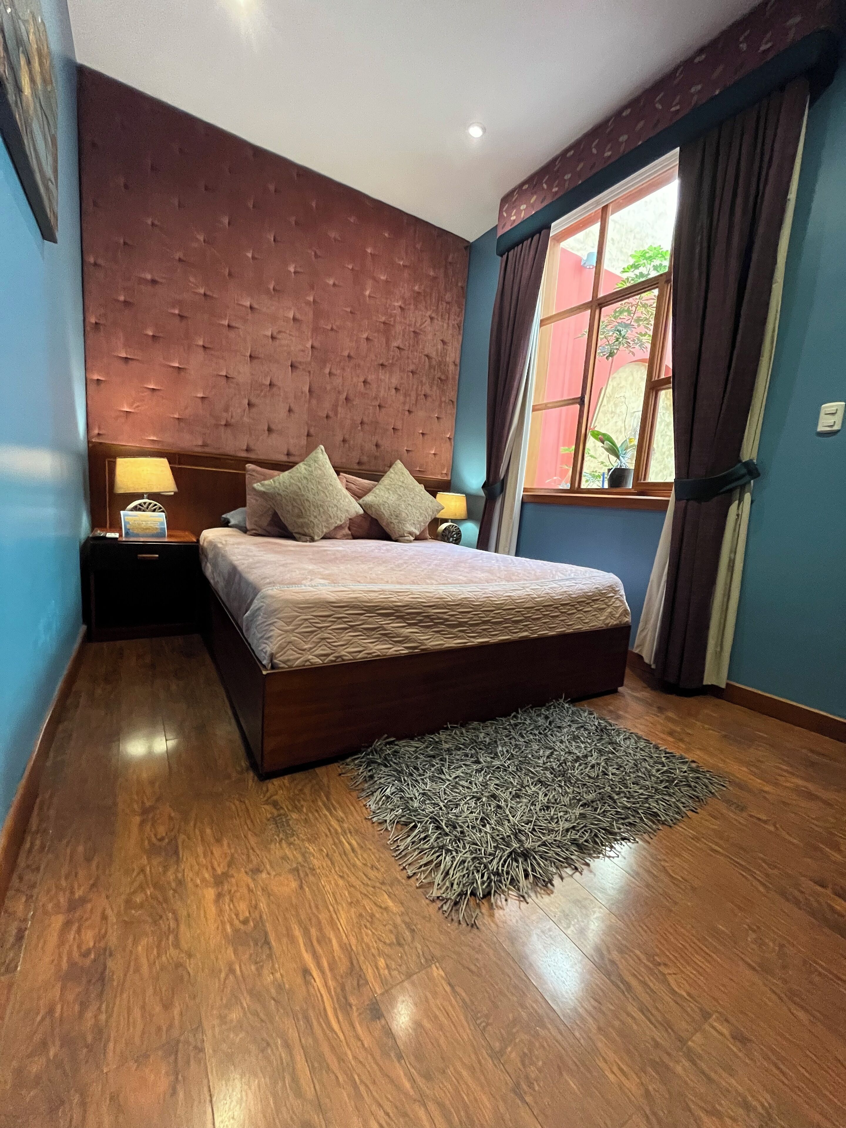 Quarto com cama grande, cabeceira de madeira, duas mesas de cabeceira com luminárias, janela com cortinas e tapete no chão.
