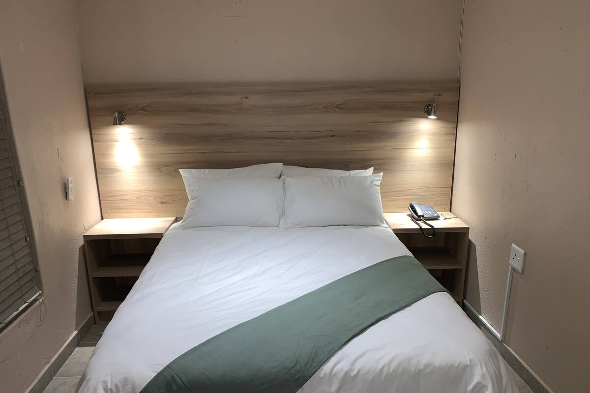 Cama bem arrumada com roupa de cama branca e verde, cabeceira de madeira, abajures e um telefone em um criado-mudo de madeira.