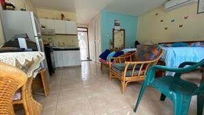 Um espaço compacto com cozinha, área de refeições e um pequeno espaço de estar com móveis de vime e uma cadeira de plástico.