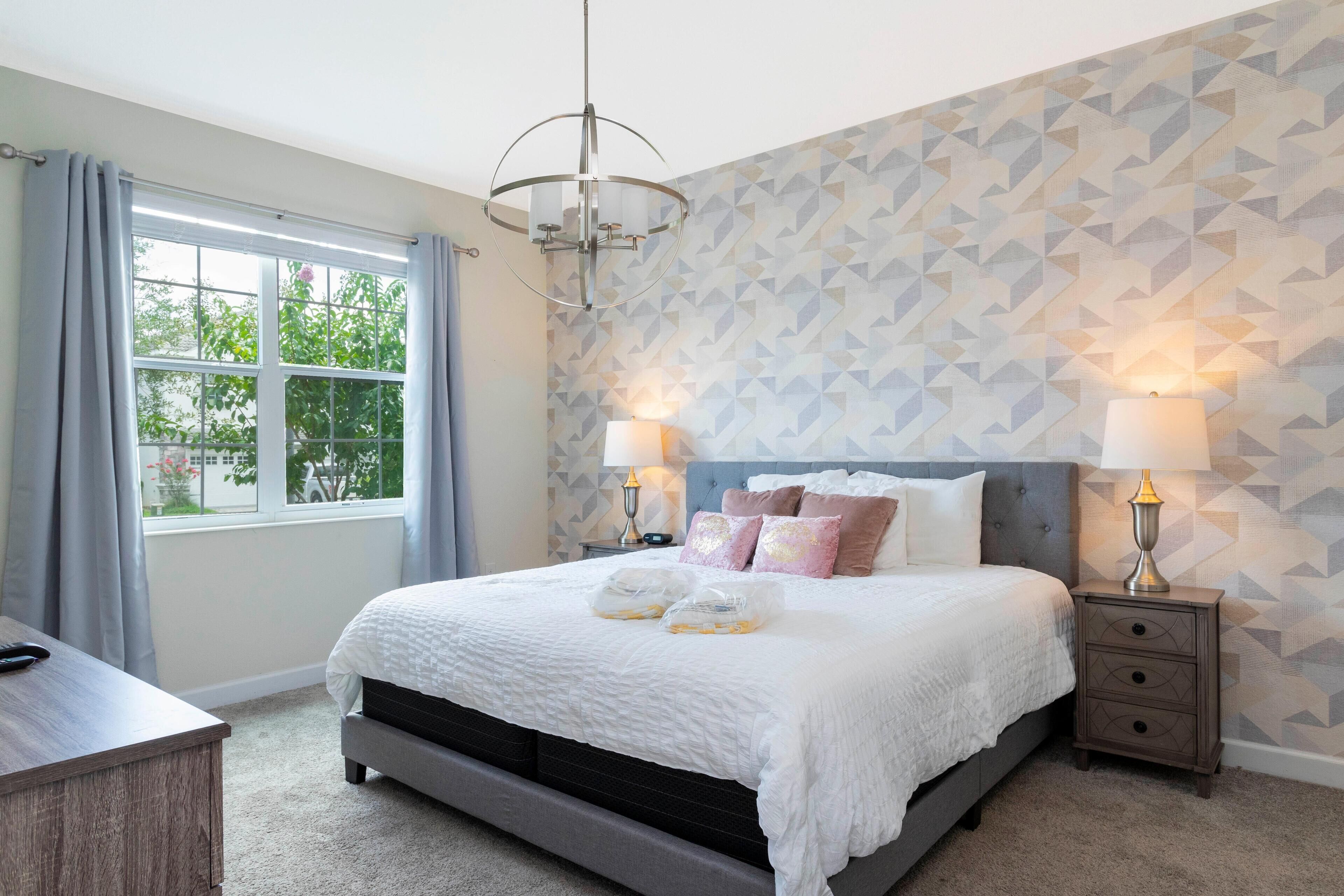 Quarto com cama grande, duas mesas de cabeceira com luminárias, janela com cortinas e parede com padrão geométrico.