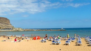 Uma praia com espreguiçadeiras e guarda-sóis, um penhasco rochoso e mar calmo.