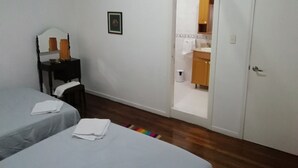 Quarto com duas camas, uma mesa de cabeceira com espelho, um quadro na parede e uma porta para o banheiro.