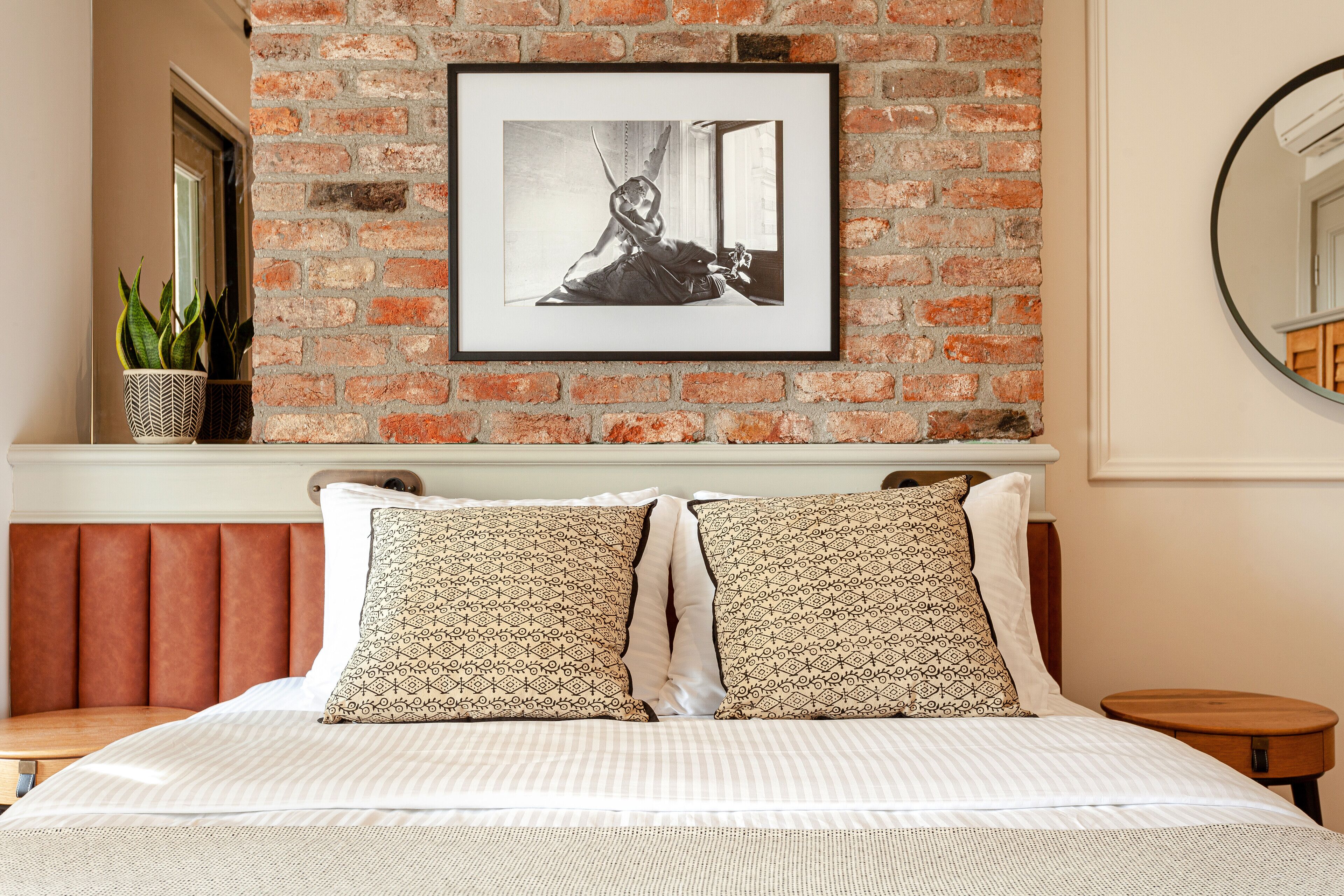 Um quarto com parede de tijolos, um quadro emoldurado, uma cama com almofadas estampadas e uma pequena mesa redonda.