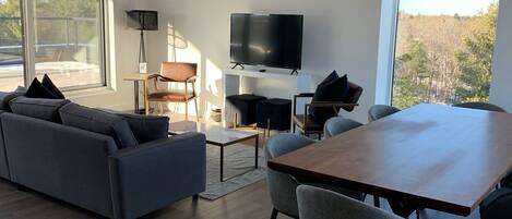 Uma sala de estar moderna com sofá, mesa de jantar, cadeiras, TV e janelas amplas com vista para árvores.
