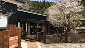 Um edifício japonês tradicional com fachada de madeira, um caminho de tijolos que leva à entrada e uma cerejeira ao fundo.