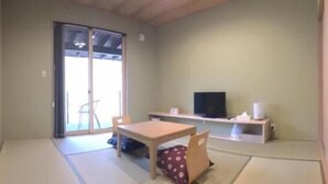 Quarto com piso de tatame, porta deslizante para área externa, televisão em suporte e pequena mesa com cadeiras.