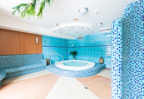Uma área interna de piscina moderna com uma banheira de hidromassagem central, azulejos de mosaico azul, bancos de madeira e um teto com iluminação embutida.