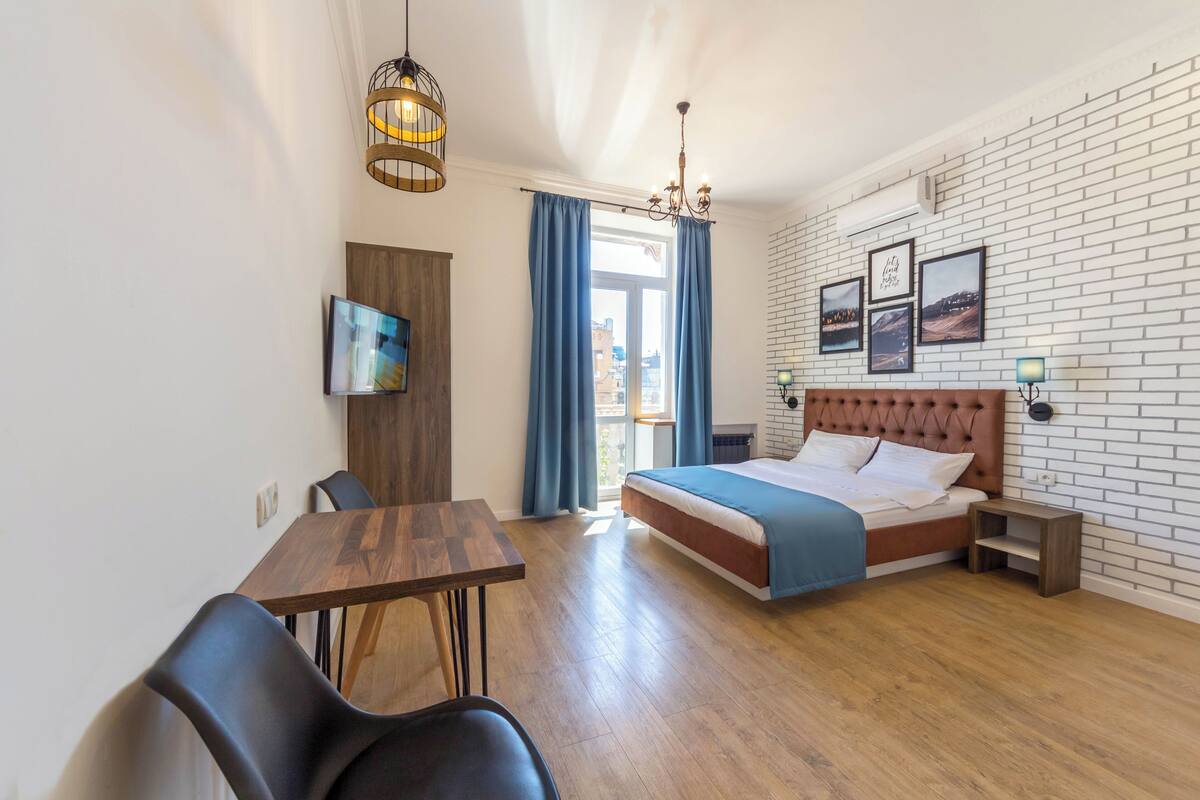 Quarto de hotel moderno com piso de madeira, cama com colcha azul, mesa de jantar com cadeiras, TV fixada na parede e lustre.