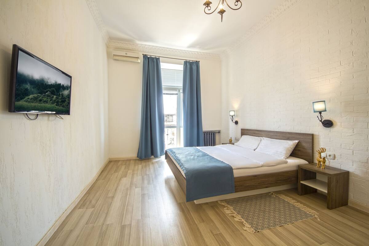 Quarto com uma cama, televisão, mesa de cabeceira e janela com cortinas.