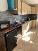 Cozinha equipada com lava-louças, fogão e micro-ondas. Há uma pia com torneira e uma máquina de café. A cozinha possui paredes revestidas de azulejos e armários de madeira.
