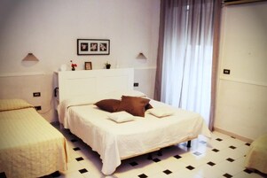 Quarto com cama, mesa de cabeceira, luminária, prateleira na parede com fotos e janela com cortinas.