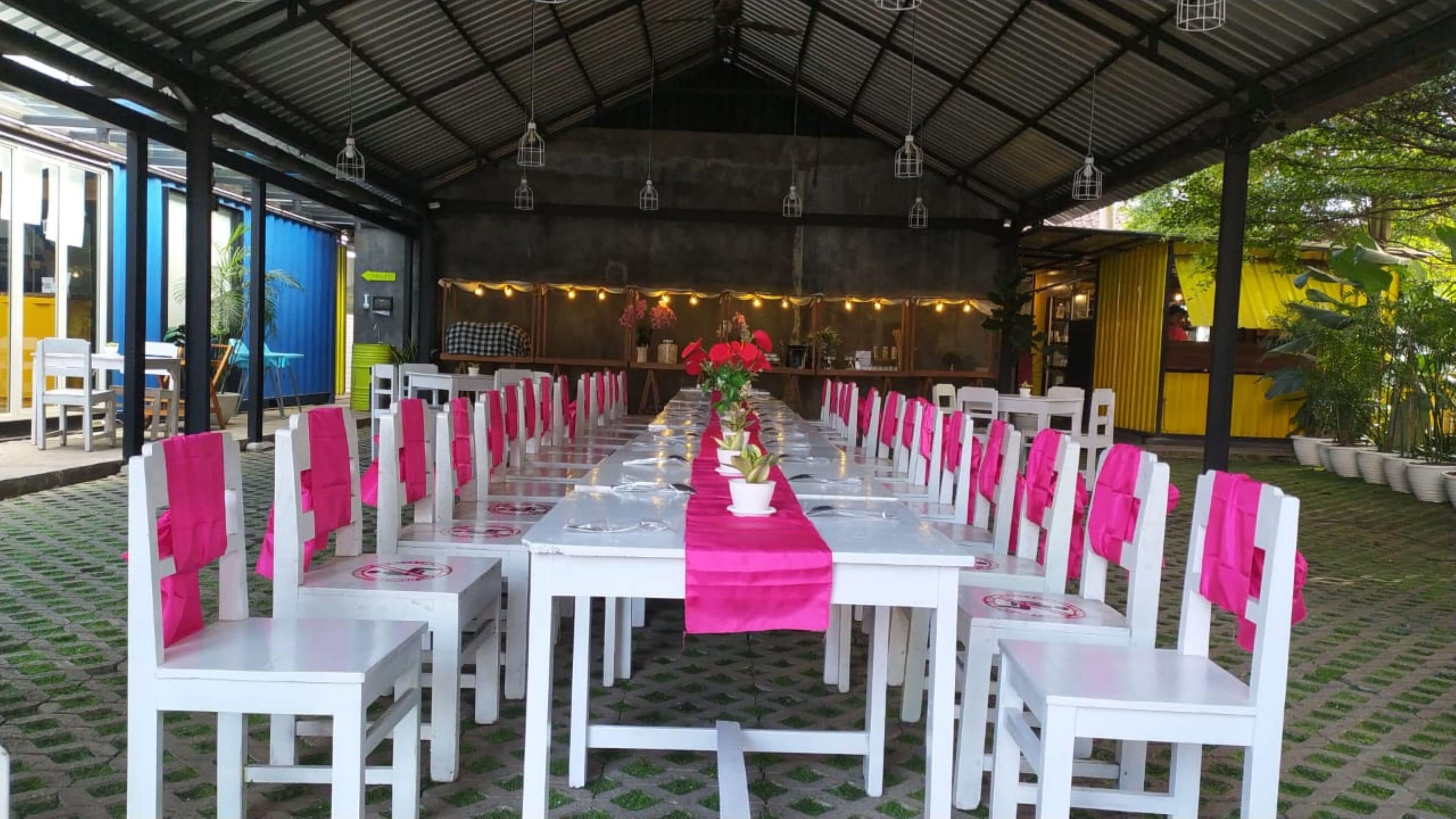 Uma área de refeições ao ar livre coberta, com mesas e cadeiras brancas, toalhas de mesa cor-de-rosa e luzes de festa.