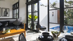Sala de estar moderna com um sofá preto, uma mesa de centro de madeira e uma mesa de jantar posta com louças pretas. Grandes janelas proporcionam vista para uma piscina e vegetação lá fora.