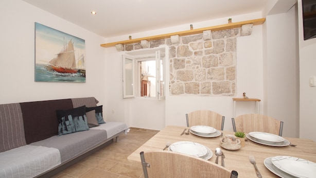 Uma área de jantar com mesa de madeira posta para quatro pessoas, um sofá com almofadas, uma parede de pedra e um quadro de um veleiro.