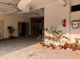 Um espaço amplo e coberto, com piso de azulejos, um suporte de metal para plantas e um extintor de incêndio fixado na parede.