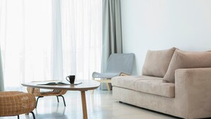 Uma sala de estar moderna com um sofá bege, uma mesa de madeira redonda com um livro e uma xícara, e uma cadeira de vime.