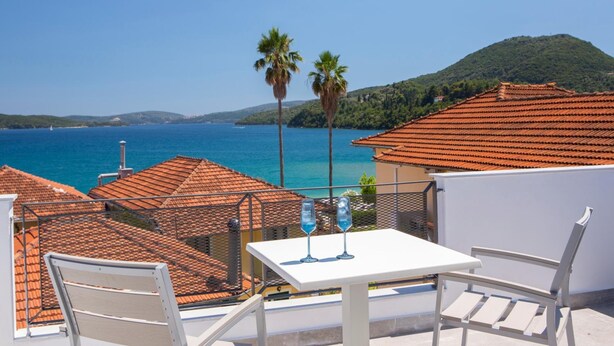 Terraço com mesa e cadeiras brancas, com vista para a costa, telhados de barro avermelhados e palmeiras.