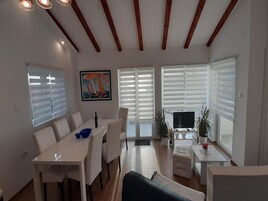 Uma sala de estar moderna com área de jantar, janelas amplas com persianas, um quadro na parede e uma TV em um móvel.