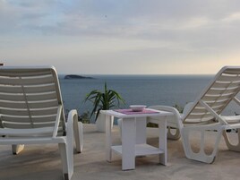 Um terraço à beira-mar com duas espreguiçadeiras brancas, uma mesinha e um vaso de plantas.