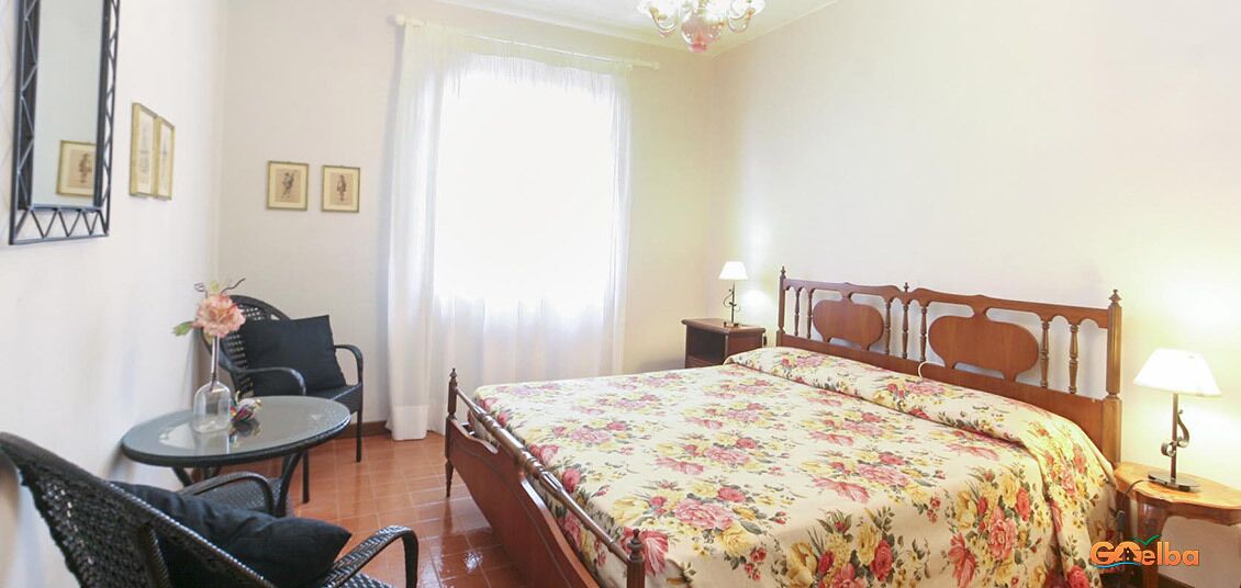Quarto com cama de madeira, uma mesa redonda pequena, uma cadeira e uma mesinha de cabeceira com abajur.
