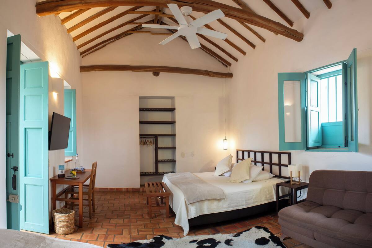 Quarto com cama, mesa de cabeceira, sofá, teto de madeira, janela com persianas azuis e piso de azulejos.