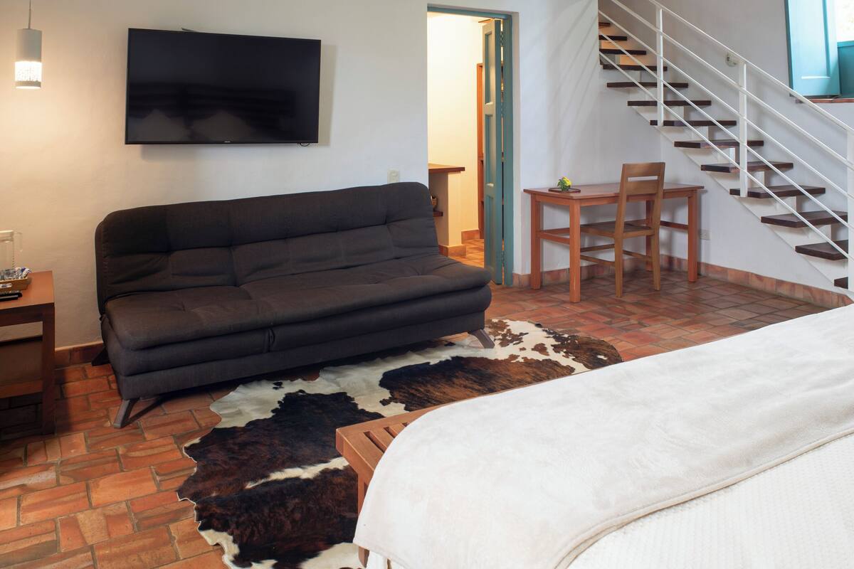 Um quarto espaçoso com teto alto, um sofá grande, uma mesa de madeira e uma escada que leva a um andar superior.