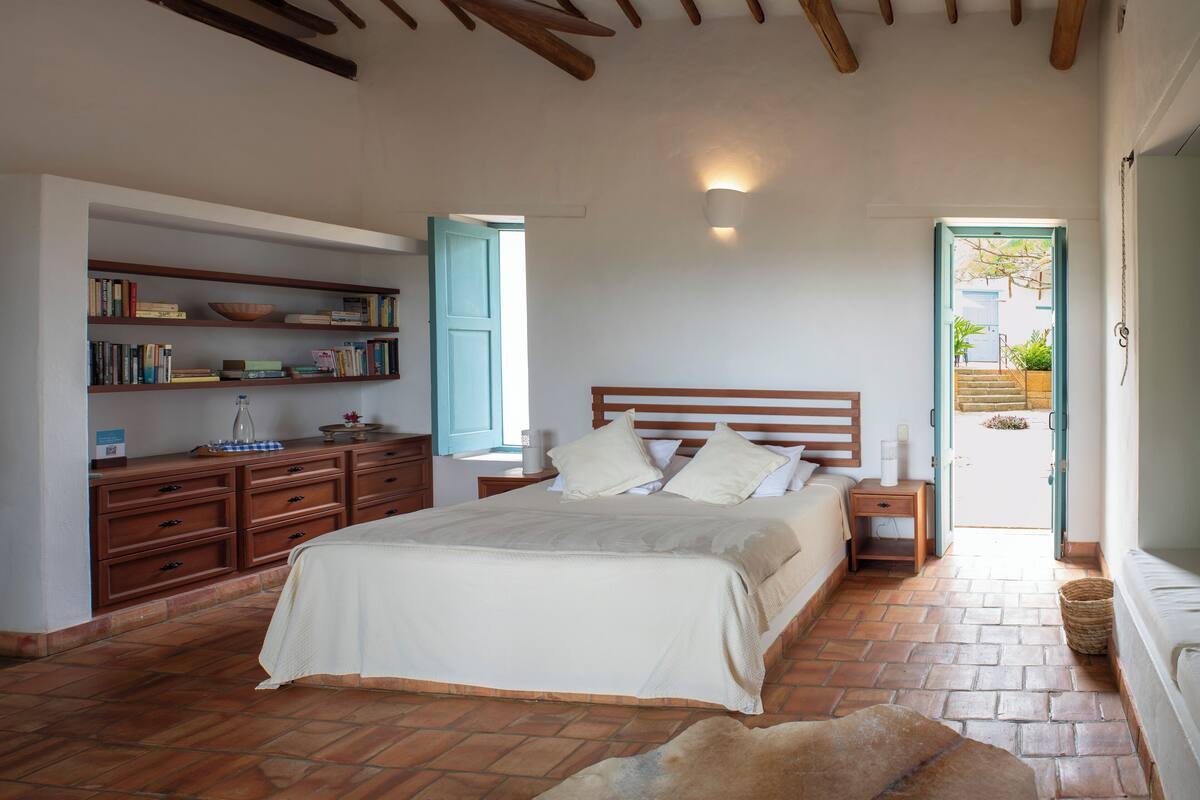Quarto com cama de madeira, mesa de cabeceira, estante e vista para o exterior.