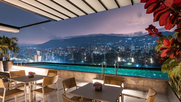 Área externa com mesas e cadeiras, vista para o horizonte da cidade à noite, piscina e plantas em vasos.