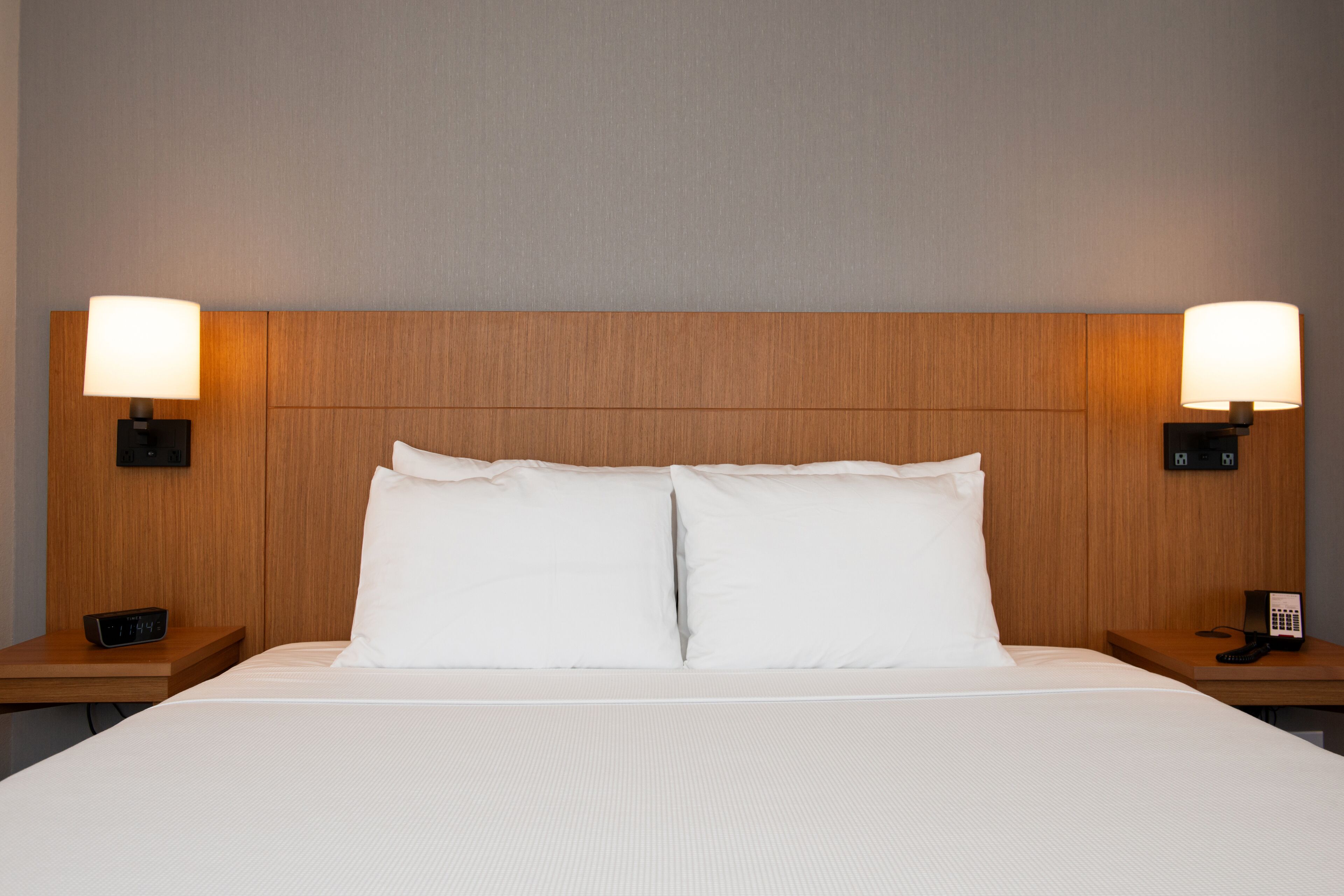 Quarto de hotel com uma cama grande, duas mesas de cabeceira, um painel de cama de madeira e duas luminárias fixadas na parede.