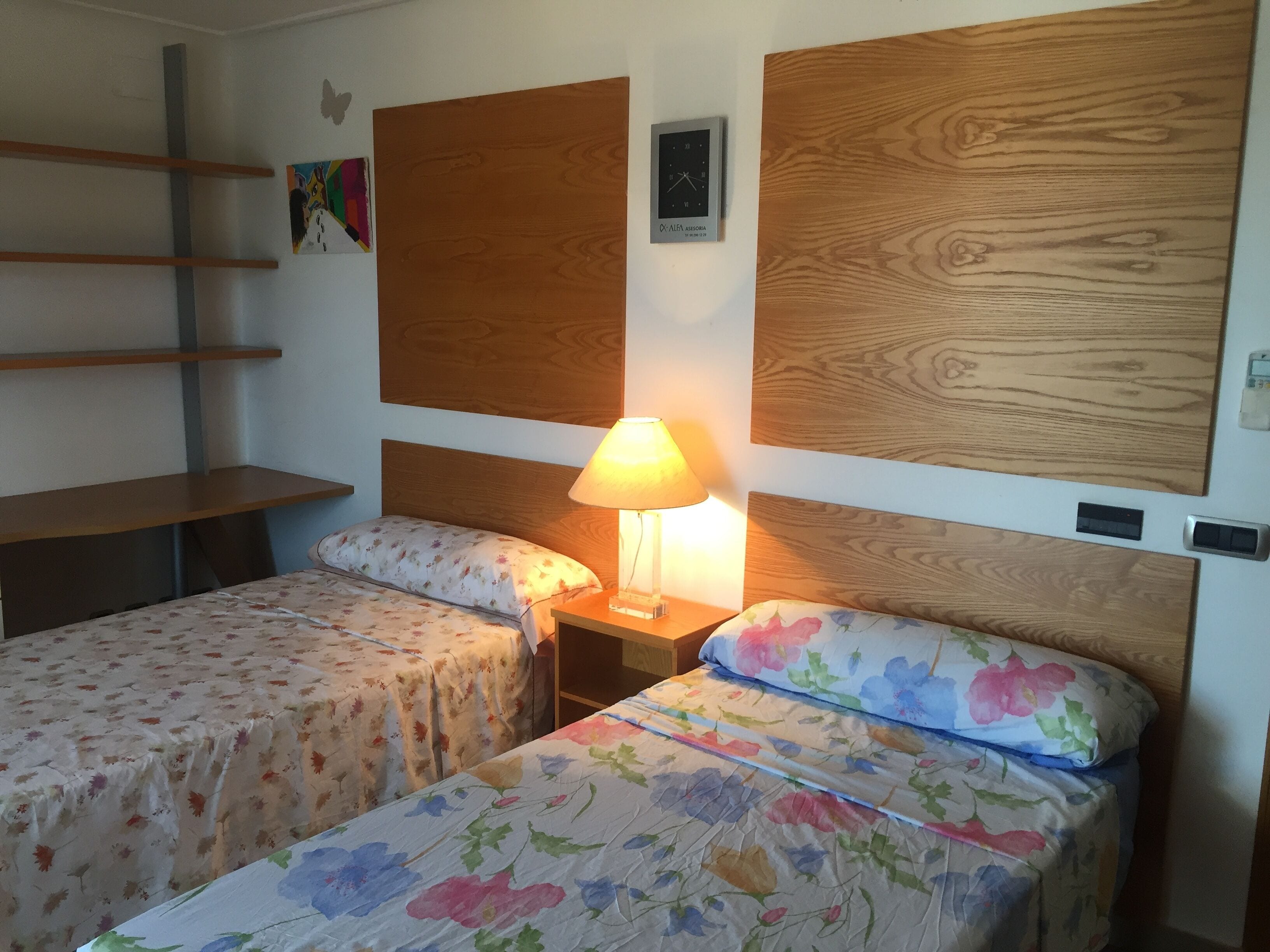 Quarto com duas camas de solteiro, mesa de cabeceira com luminária, cabeceiras de madeira, relógio de parede e decoração de borboleta.
