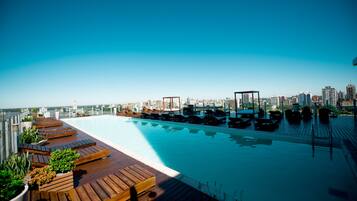 Piscina infinita no terraço, com espreguiçadeiras de madeira e plantas em vasos, com vista para o horizonte urbano.