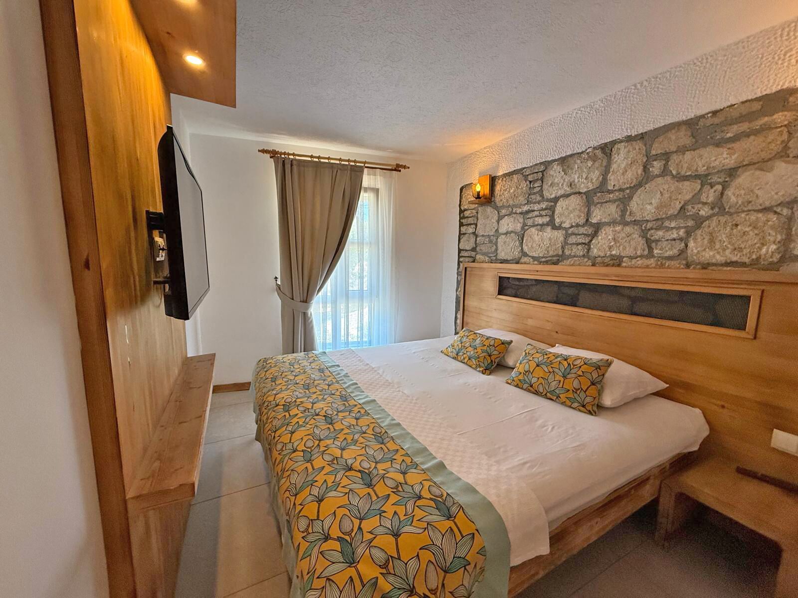 Quarto com cabeceira de madeira, parede de pedra, cama com roupa de cama estampada, um banco de madeira e uma janela com cortinas.