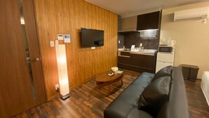 Quarto de hotel compacto com guarda-roupa de madeira, televisão de ecrã plano na parede, kitchenette com micro-ondas e sofá.