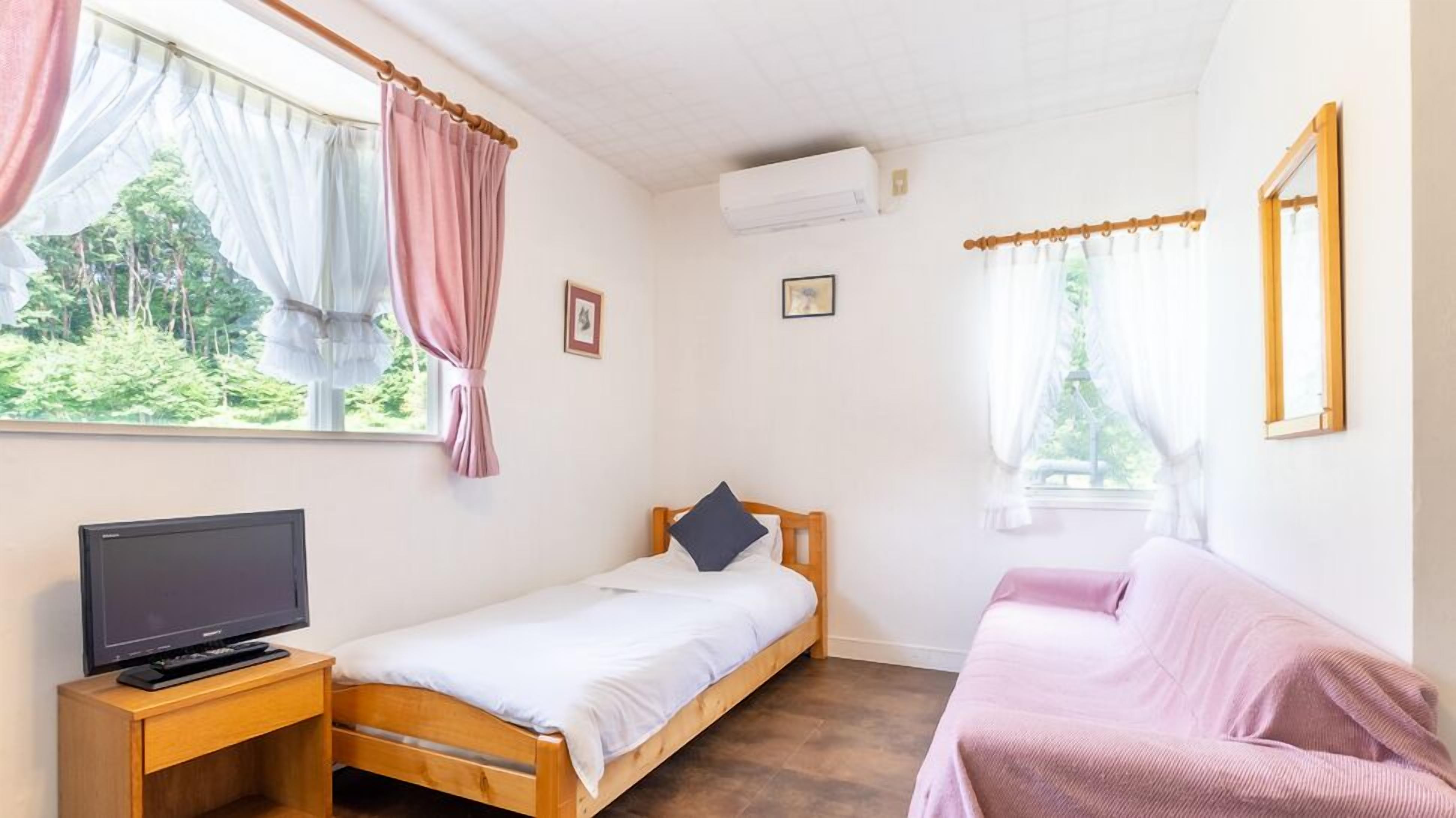 Quarto com cama, televisão em suporte de madeira, sofá rosa e duas janelas com cortinas.