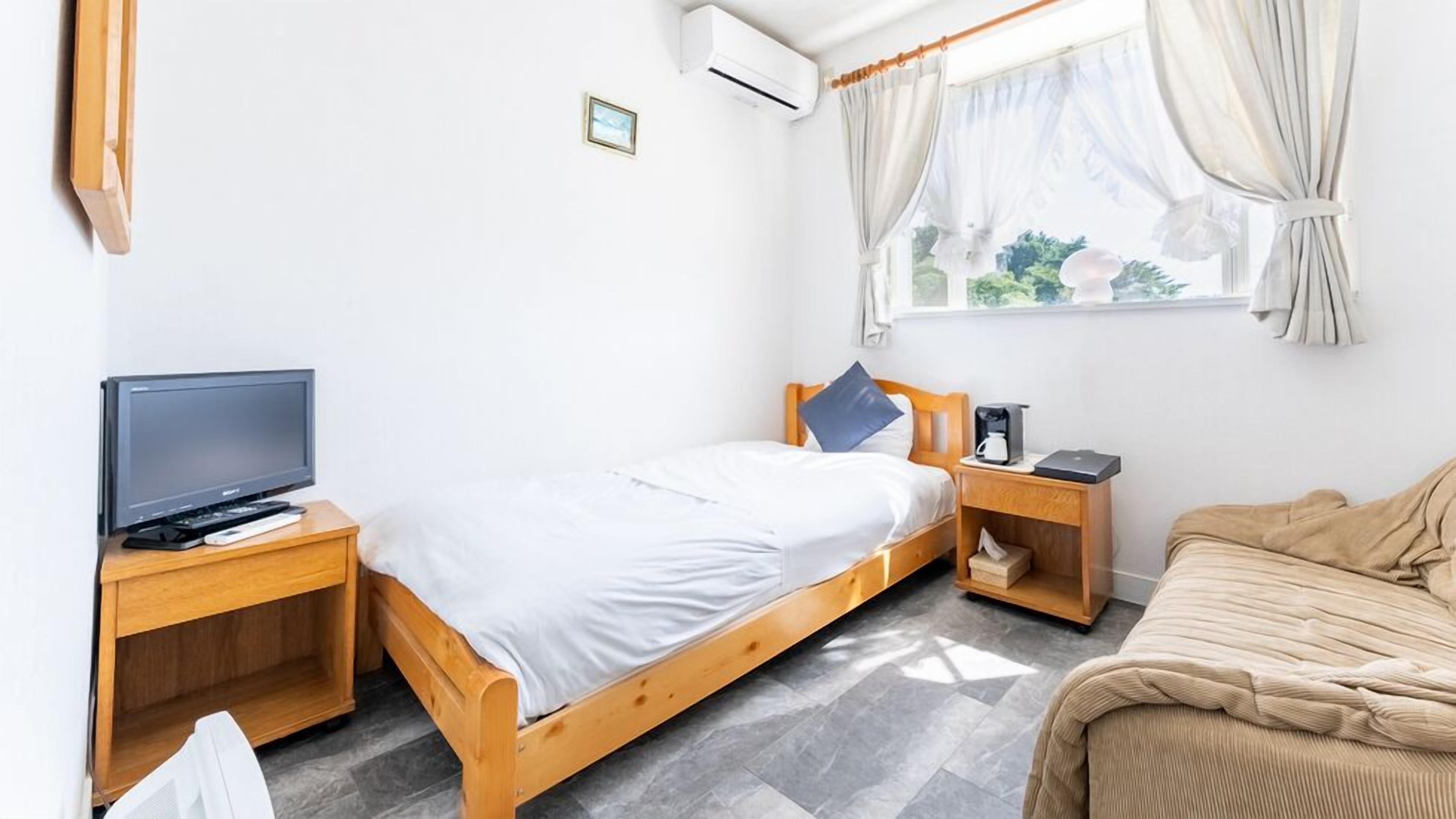 Um quarto com uma cama, uma televisão sobre uma mesa de cabeceira de madeira, uma mesinha de cabeceira com uma luminária, uma janela com cortinas e um sofá.