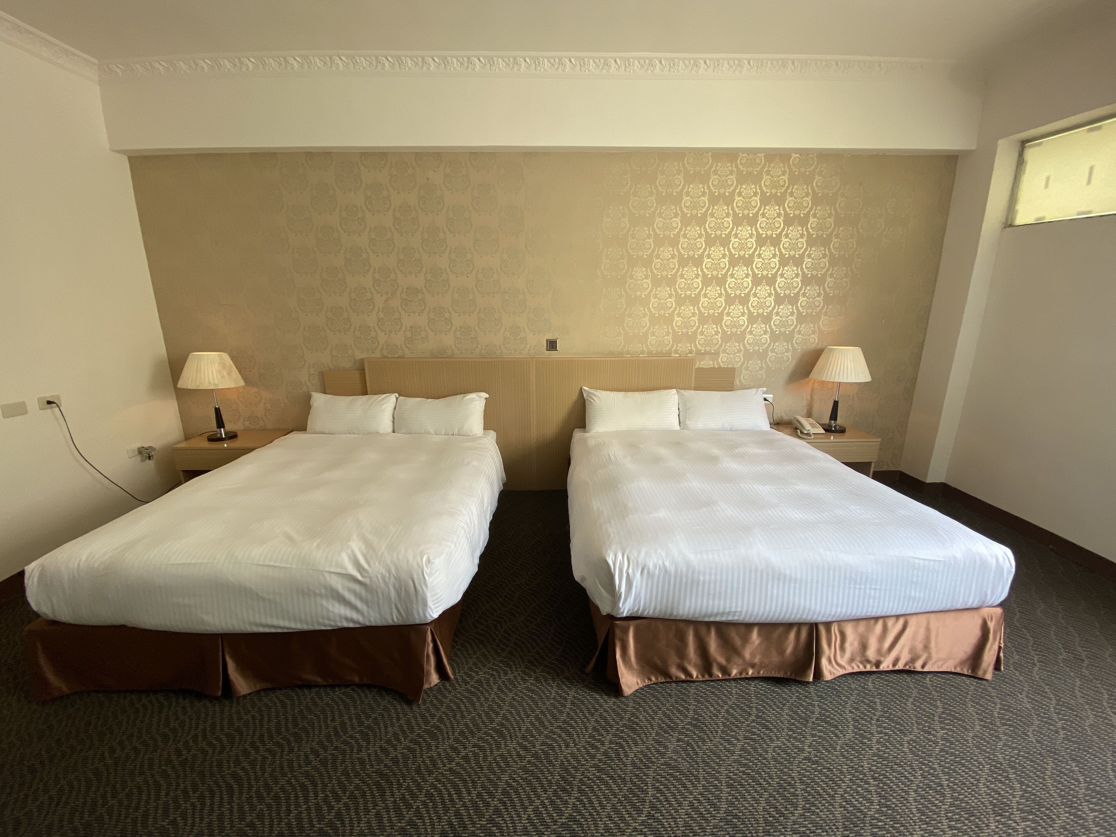Quarto de hotel com duas camas, papel de parede estampado, piso coberto por carpete e luminárias de cabeceira.