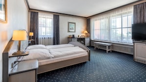 Quarto de hotel com uma cama de casal, duas mesas de cabeceira com luminárias, uma escrivaninha com cadeira, uma televisão e uma janela grande com cortinas.