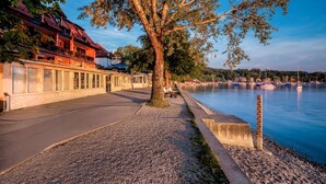 Hotel na orla marítima com fileira de quartos, calçadão arborizado e barcos ao fundo.
