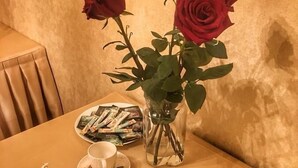 Uma mesa posta com xícaras e pires brancos, um vaso com rosas vermelhas e uma bandeja com revistas.