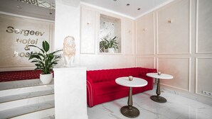 Um saguão de hotel com um sofá vermelho, duas mesinhas redondas, uma planta em vaso e uma estátua decorativa de leão.