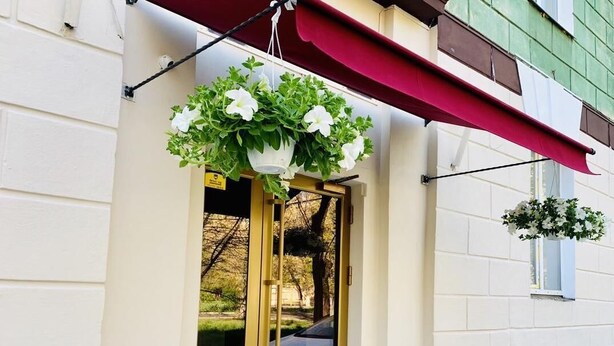 Entrada de hotel com porta de vidro, arranjo de flores pendente e toldo vermelho.