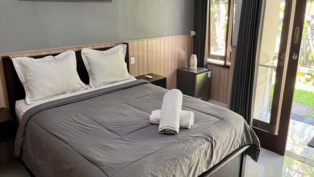 Uma cama bem arrumada com um edredom cinza e duas almofadas brancas, uma mesa de cabeceira com uma luminária e vista para um jardim através de uma porta de vidro.