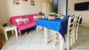 Uma área de refeições com uma mesa coberta por uma toalha azul, rodeada por cadeiras brancas. Um sofá rosa com almofadas listradas está encostado na parede, e uma pequena mesa com um livro e uma xícara são visíveis.