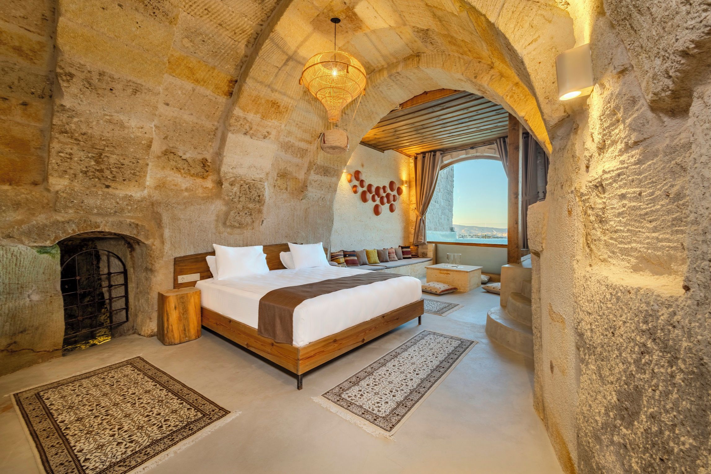 Quarto com parede de pedra, cama, cabeceira de madeira, luminária pendente e vista pela janela.