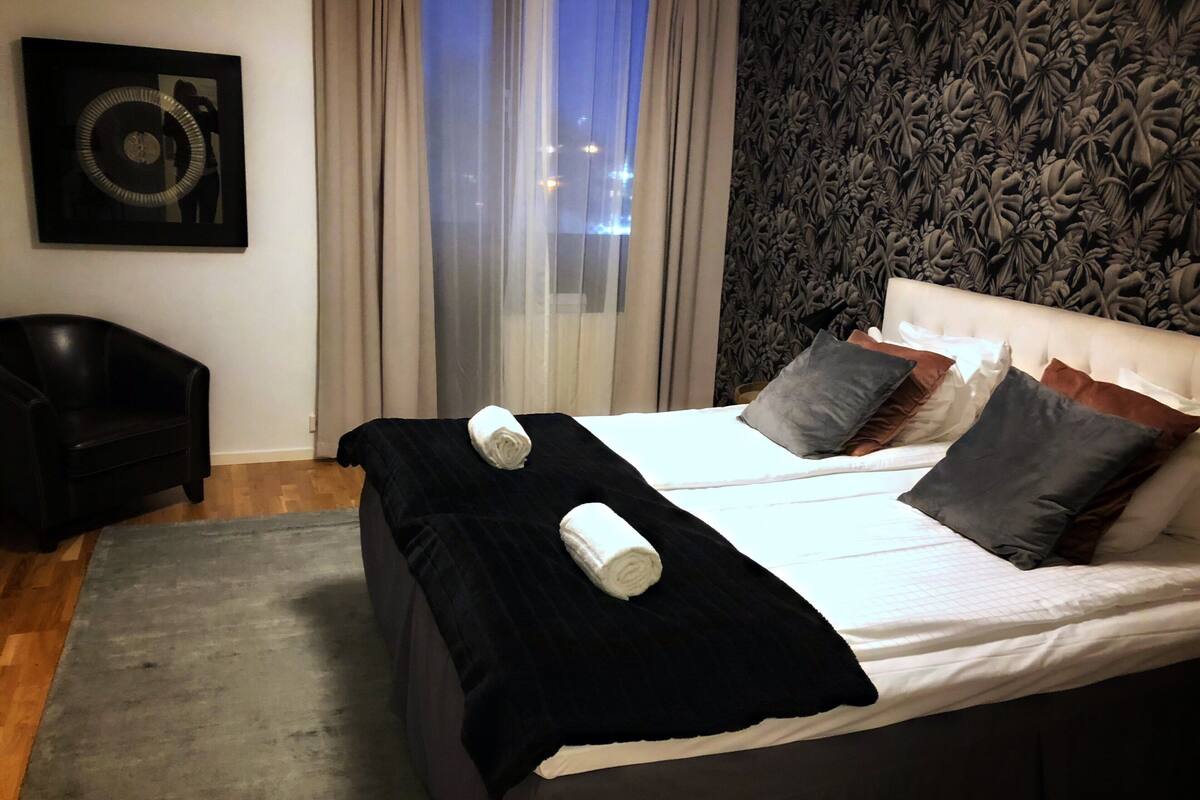 Quarto de hotel com cama, poltrona preta, janela com cortinas e parede com padrão floral.