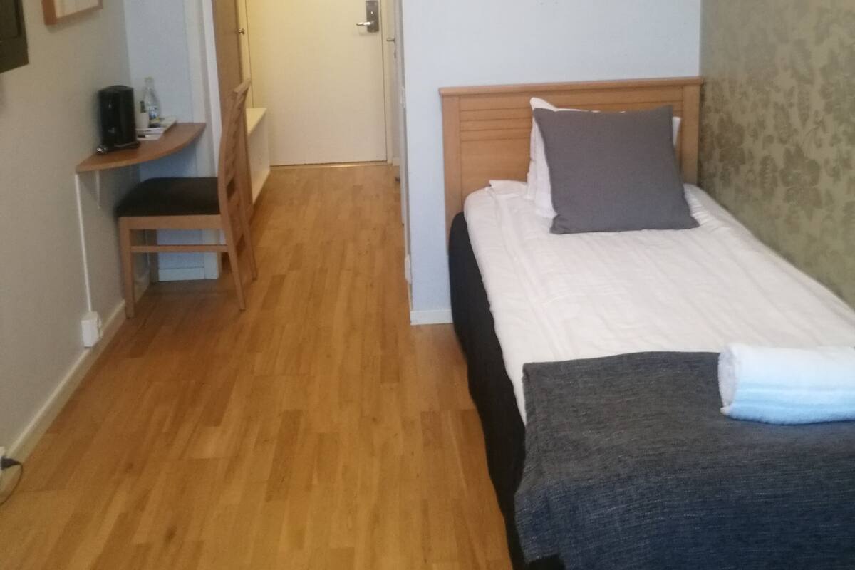 Quarto com piso de madeira, uma cama com edredom branco e travesseiro cinza, mesa de cabeceira de madeira e uma cadeira.
