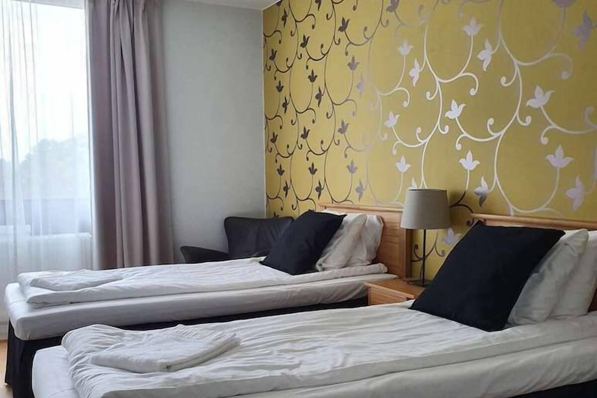 Quarto de hotel com duas camas de solteiro, mesa de cabeceira com luminária e janela com cortinas.