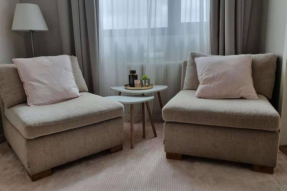 Um quarto com duas poltronas bege, uma mesa redonda pequena com uma luminária e cortinas leves.